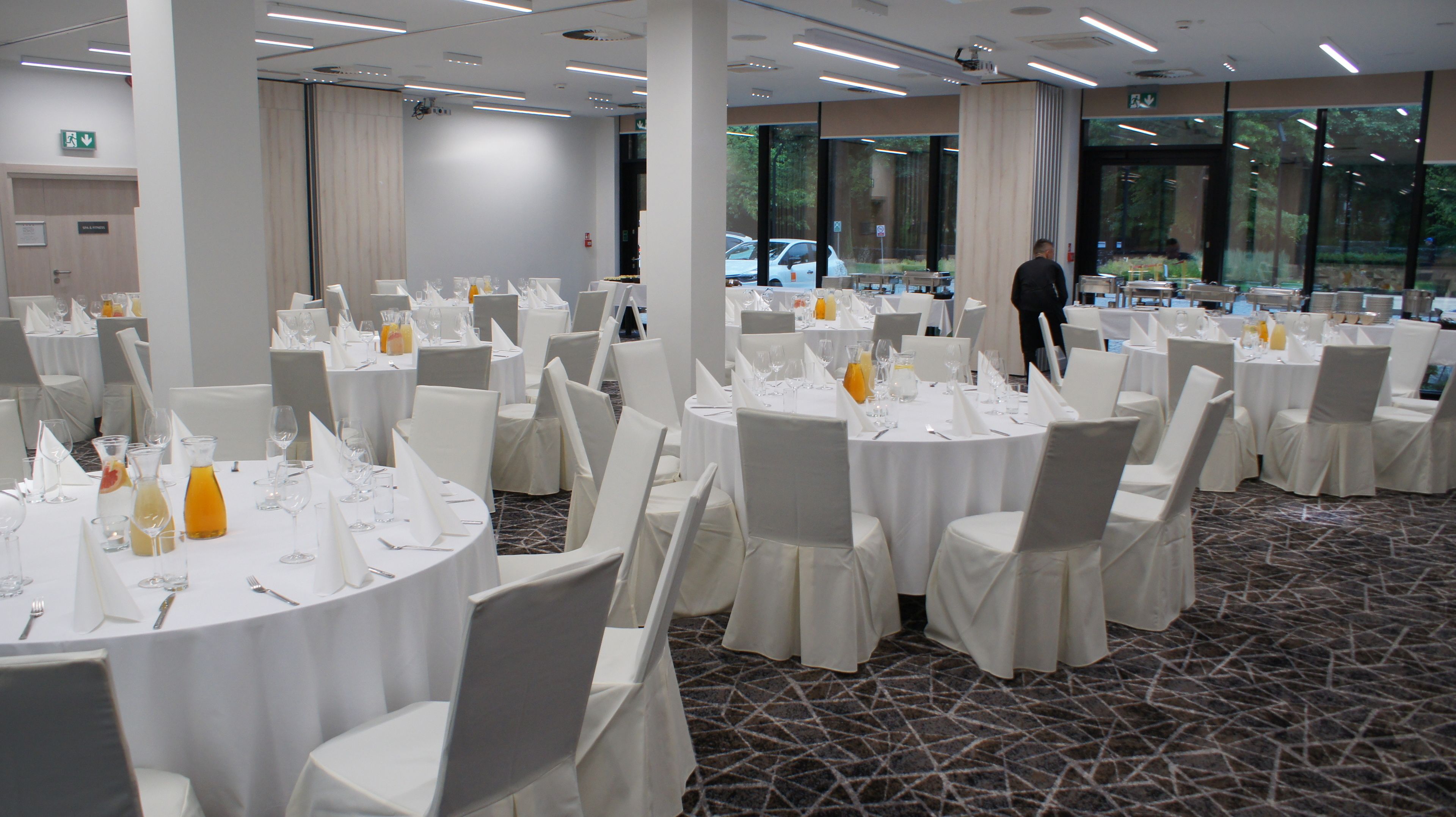 Uma sala de banquetes com mesas redondas preparadas para refeições, cadeiras brancas e piso coberto de carpete.