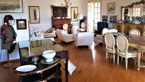 Uma sala de estar ampla com área de jantar, destacando uma mesa de centro de vidro, sofás brancos e uma mesa de jantar de madeira com cadeiras.