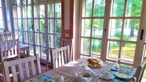 Uma sala de jantar com mesa posta para refeição, rodeada de cadeiras, e janelas amplas com vista para o verde lá fora.