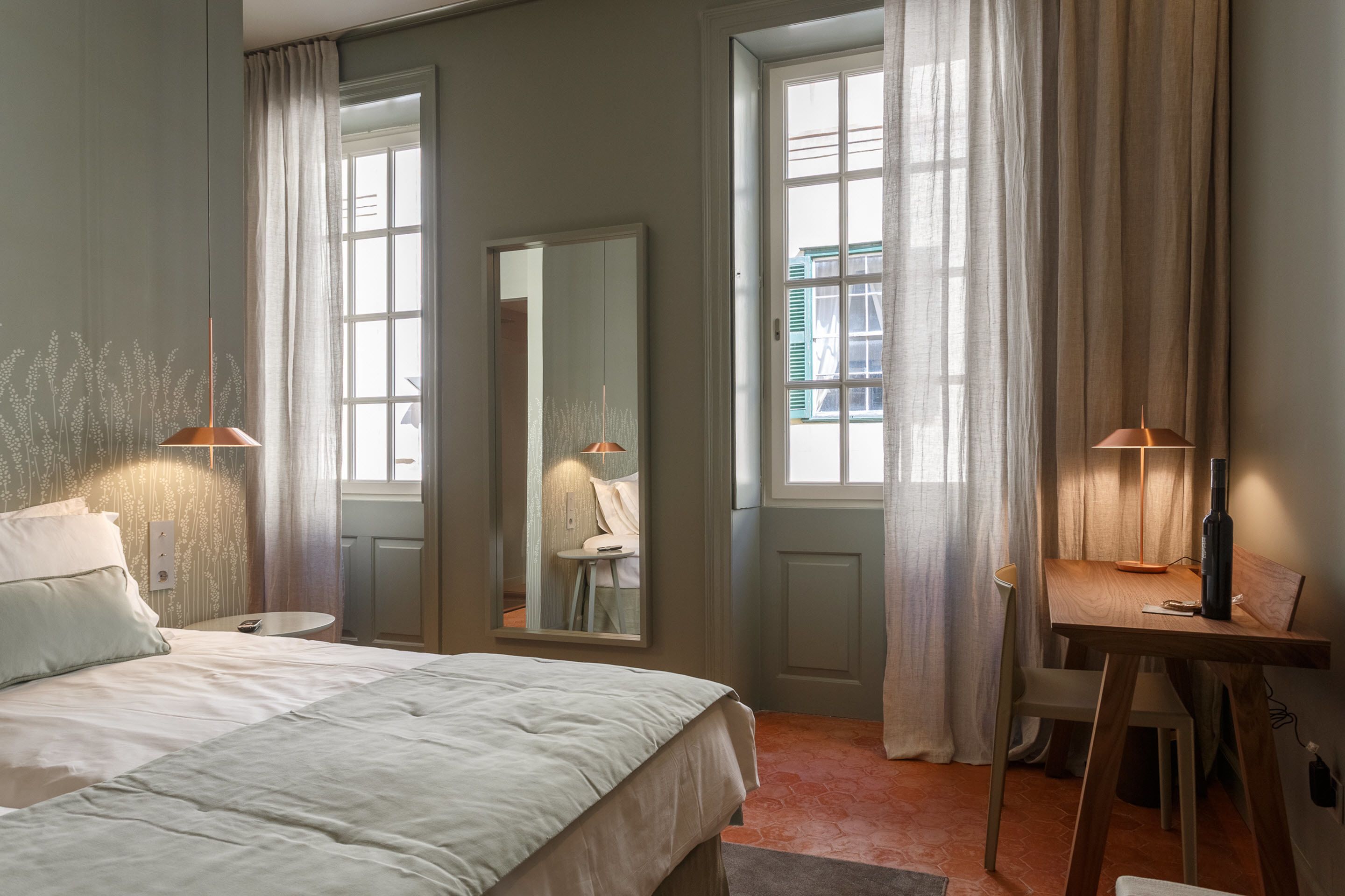 Quarto com cama, escrivaninha, cadeira, espelho e janela com cortinas.