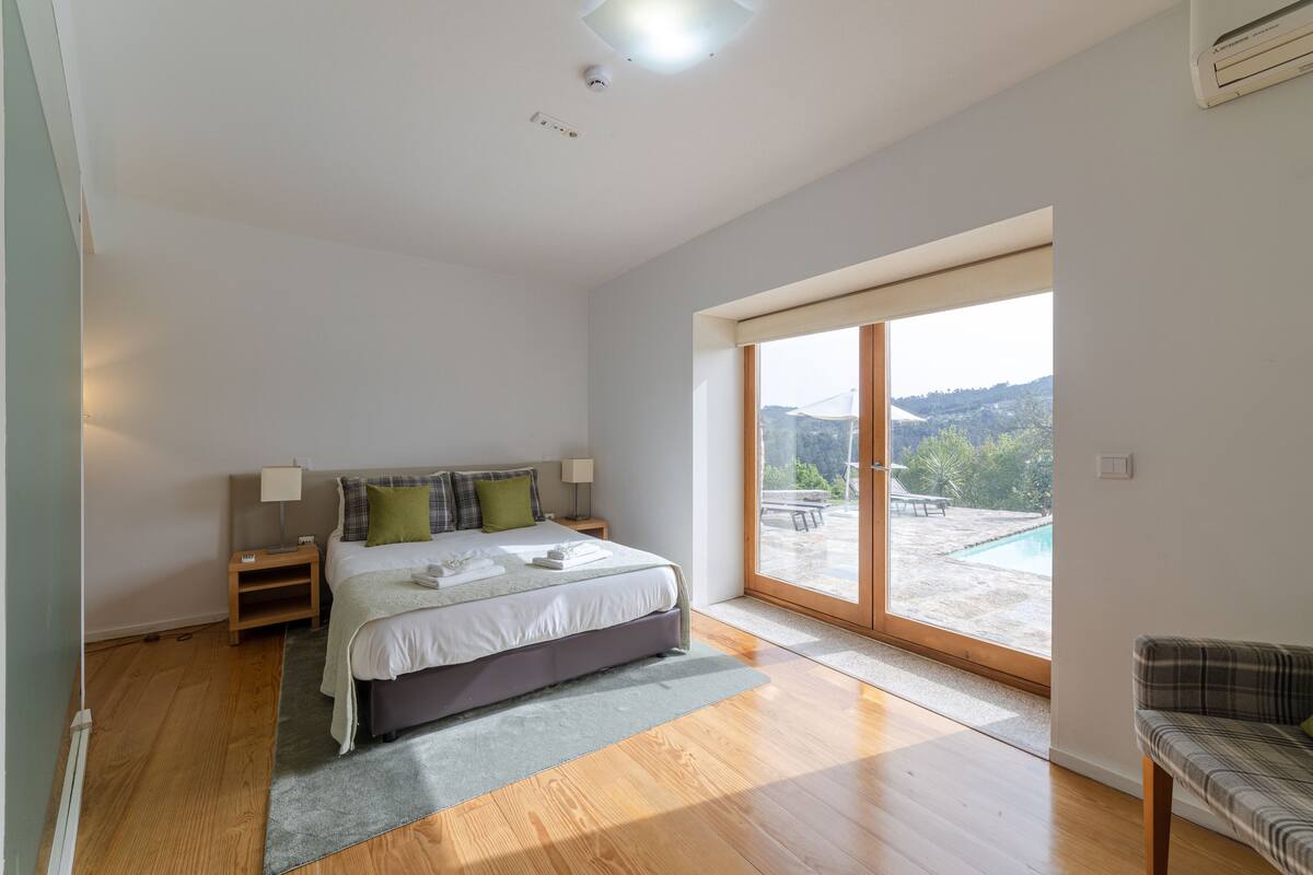 Quarto com cama, mesas de cabeceira, um sofá e uma grande porta de vidro deslizante que dá para uma varanda com piscina.