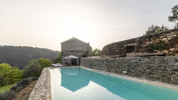 Uma piscina de paredes de pedra com superfície de água azul cristalina, rodeada por vegetação exuberante e um edifício de pedra ao fundo.