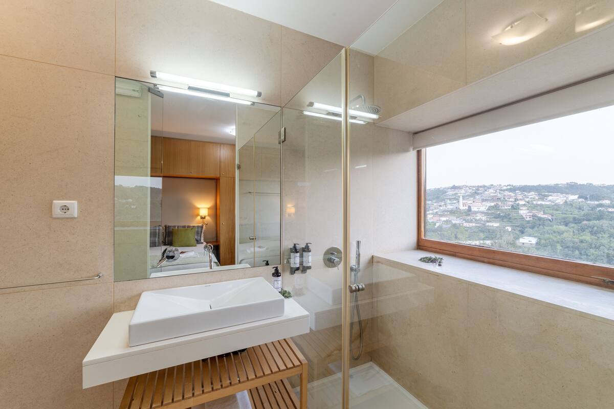 Uma casa de banho moderna com chuveiro em cabine de vidro, um espelho grande e vista para a cidade através de uma janela.