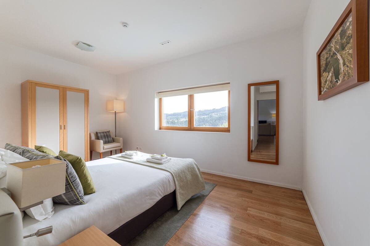 Quarto de hotel moderno com uma cama grande, uma escrivaninha, uma cadeira, um abajur e uma janela com vista.
