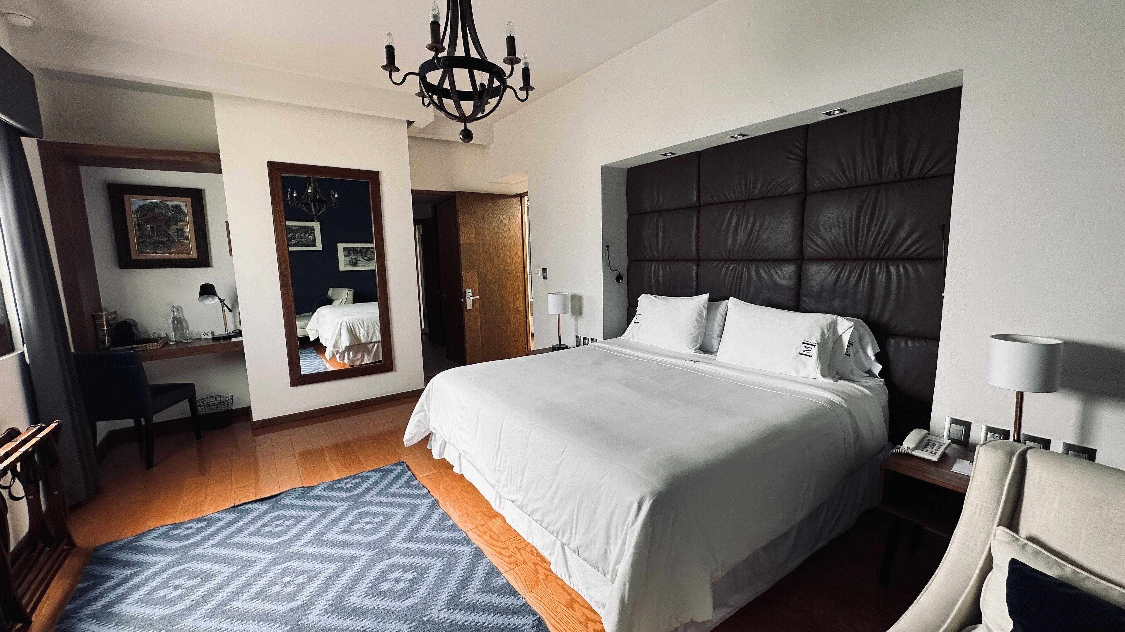 Quarto de hotel moderno com uma cama grande, cabeceira escura, escrivaninha com cadeira, luminária de mesa e um espelho.