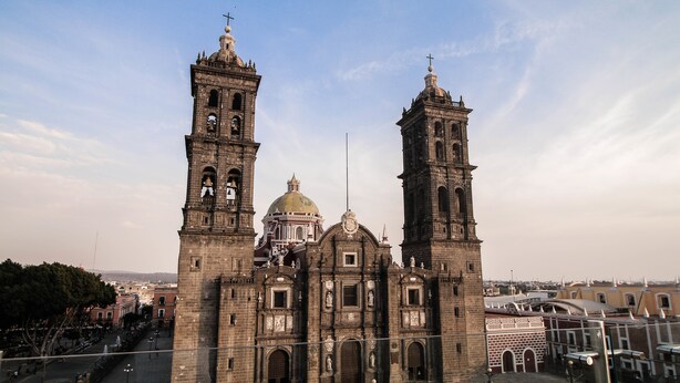 Uma catedral histórica com duas torres imponentes e uma cúpula central, rodeada por uma praça com algumas pessoas e veículos.