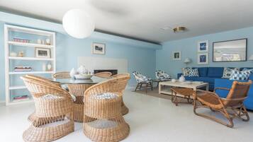 Uma sala de estar com uma mesa de vidro redonda cercada por cadeiras de vime, um sofá azul com almofadas estampadas e uma lareira branca.
