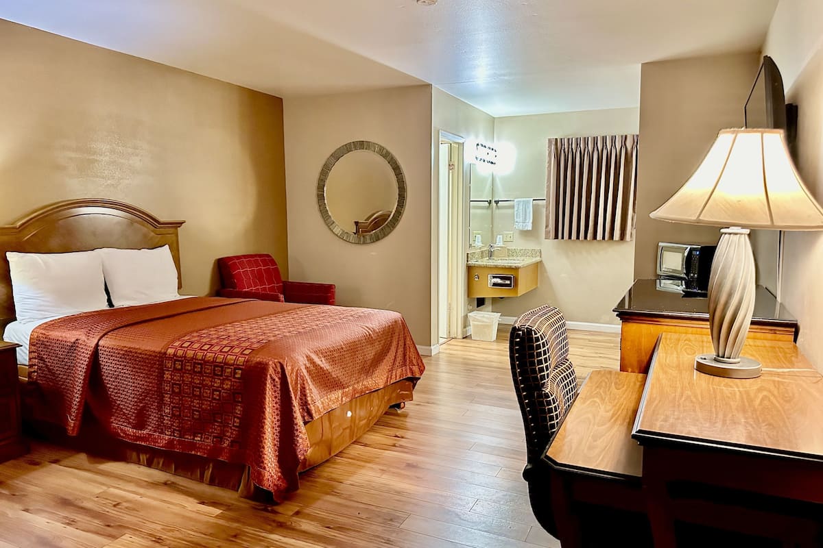 Quarto de hotel com cama de madeira, uma escrivaninha com luminária, espelho e banheiro visível por uma porta aberta.