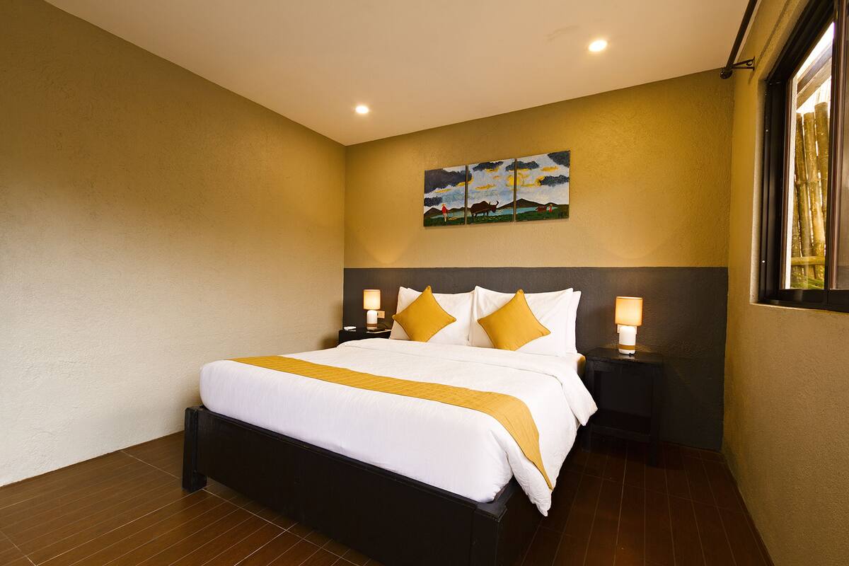 Quarto de hotel com uma cama grande, duas mesas de cabeceira com luminárias, um quadro na parede e uma janela com persianas.