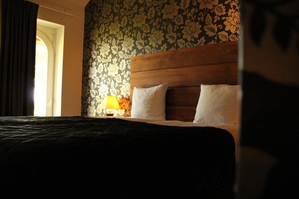 Quarto de hotel com cama, mesa de cabeceira e luminária, além de papel de parede floral.