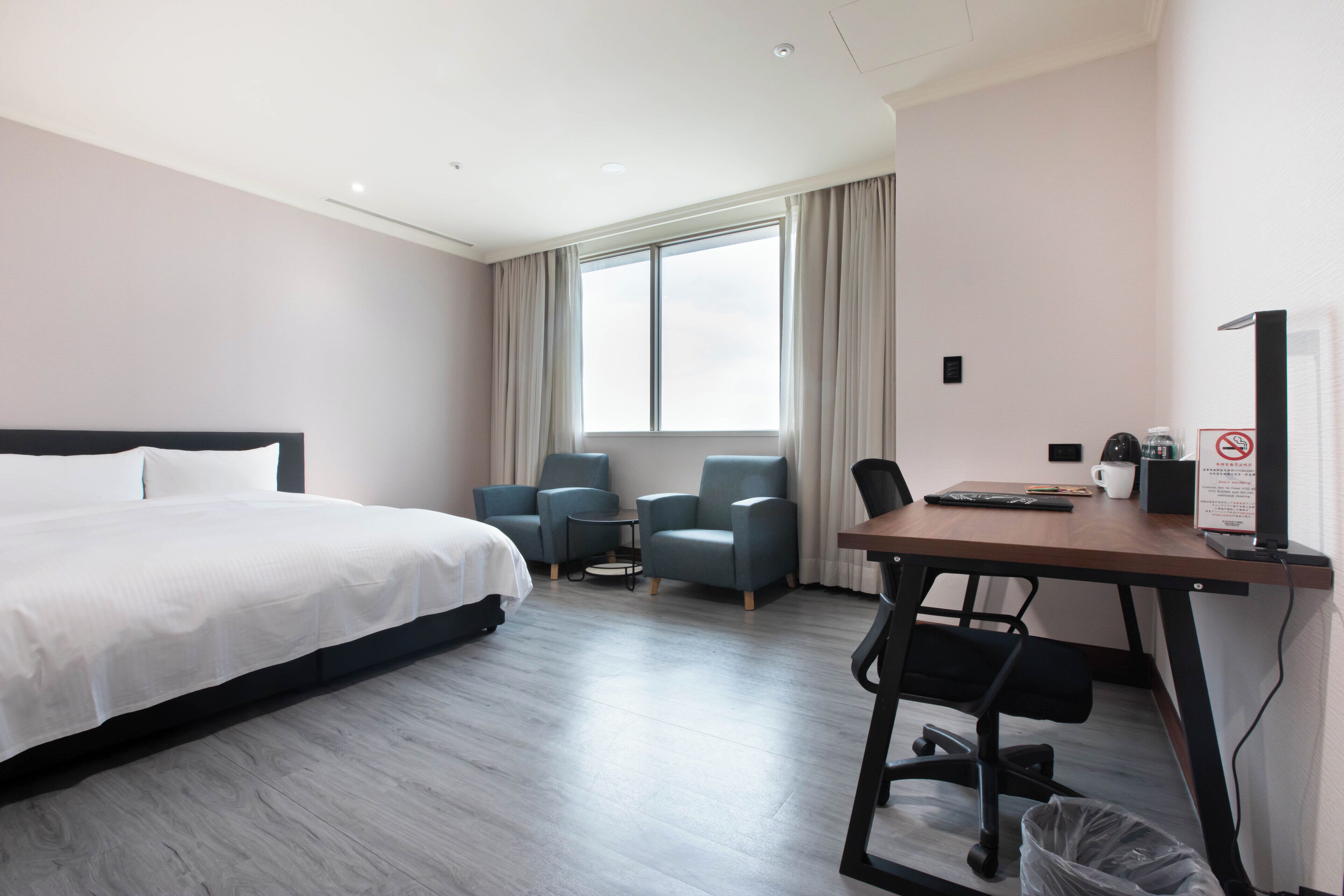 Quarto de hotel com cama, escrivaninha, duas cadeiras, mesinha e janela com cortinas.