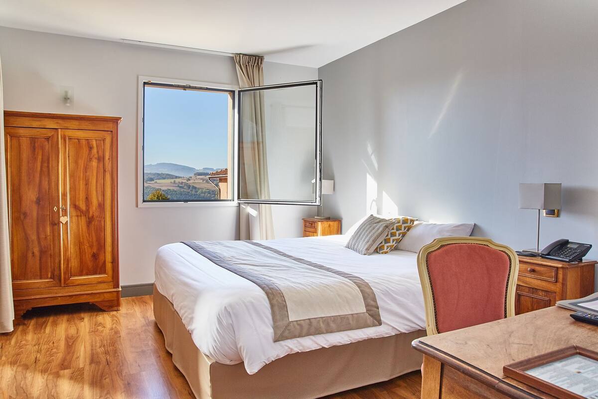 Quarto com uma cama grande, um guarda-roupa de madeira, uma escrivaninha com telefone e uma janela com vista para o campo.
