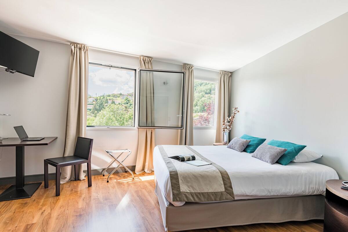 Quarto de hotel moderno com uma cama grande, uma escrivaninha com um laptop, uma cadeira e uma televisão fixada na parede.