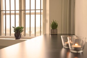 Um quarto com janela, um vaso de planta, uma vela e uma caneca listrada sobre uma mesa.