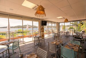 Um restaurante com mesas ao ar livre, grandes janelas e vista para a água.