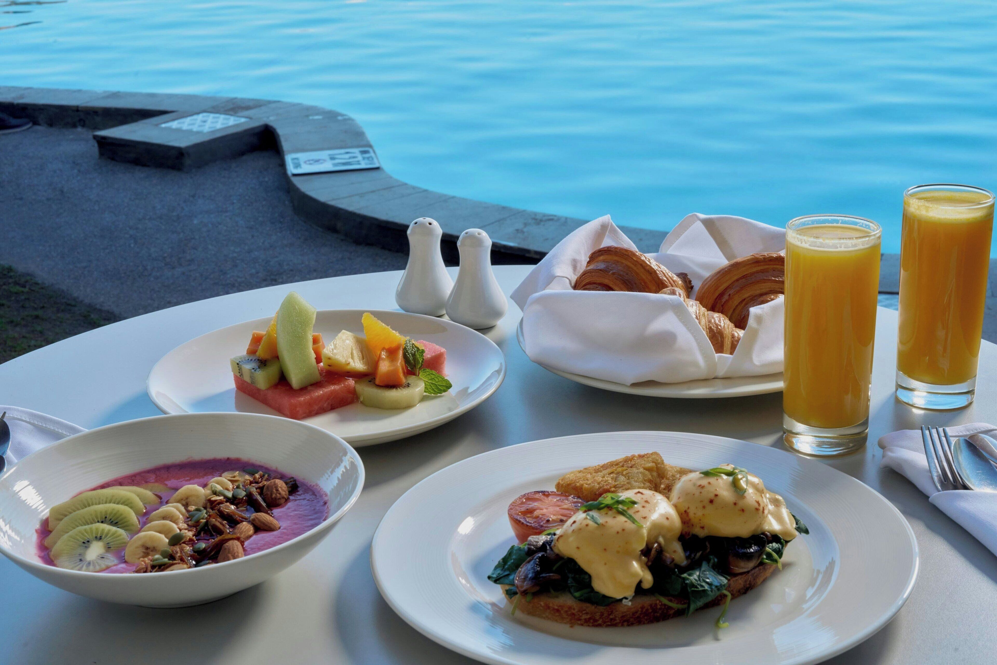 Café da manhã à beira da piscina, com croissants, frutas e suco de laranja.