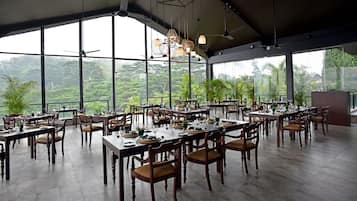 Uma ampla sala de jantar com grandes janelas que oferecem vista para vegetação exuberante. Mesas estão dispostas com cadeiras, e o ambiente apresenta um design moderno no teto.