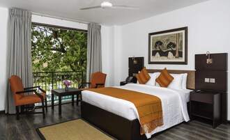 Quarto de hotel com uma cama, duas cadeiras, uma mesinha, um criado-mudo, um quadro e vista para o verde lá fora.