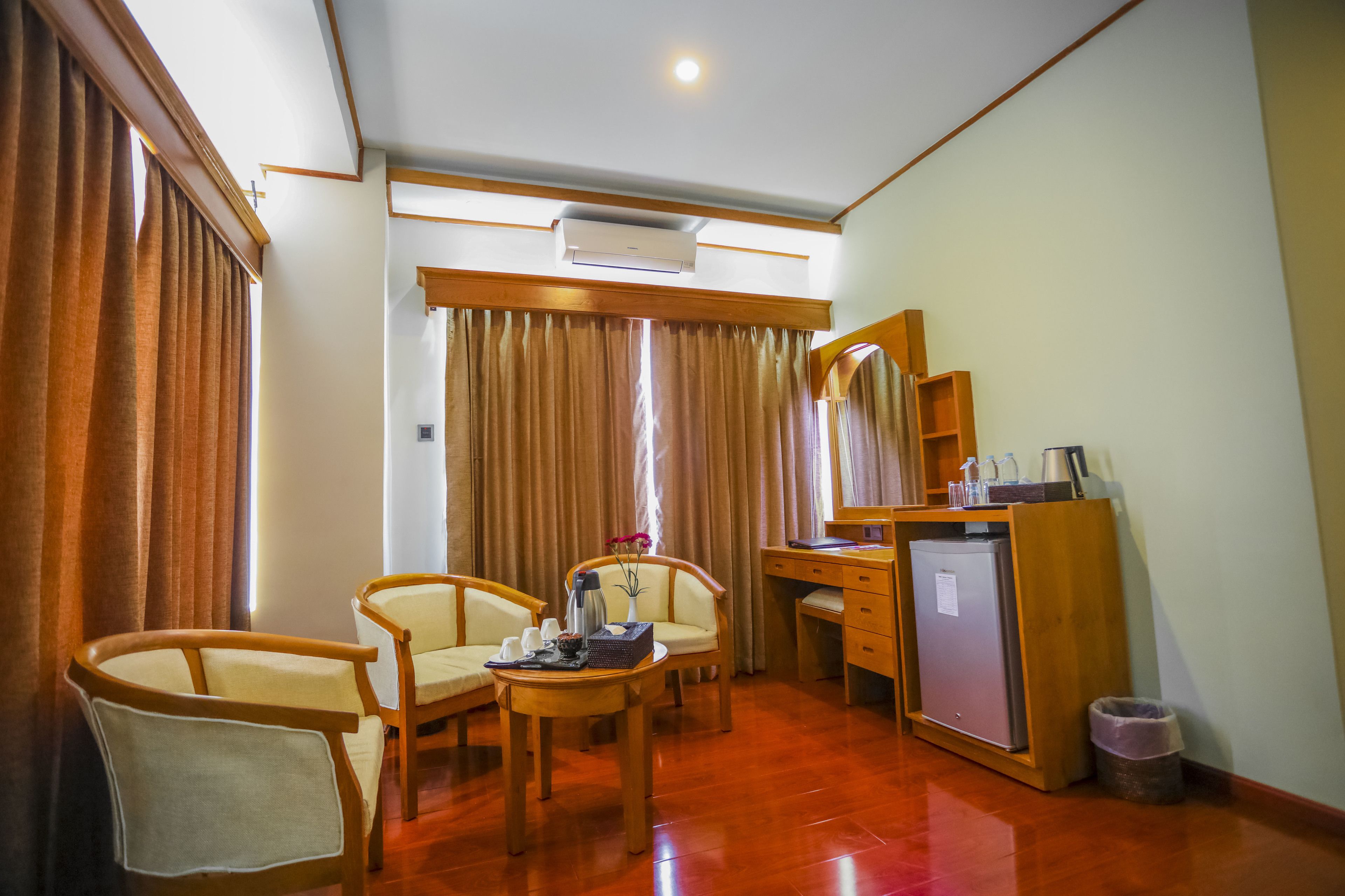 Quarto de hotel com escrivaninha de madeira, duas cadeiras, um pequeno frigorífico e um cesto de lixo.