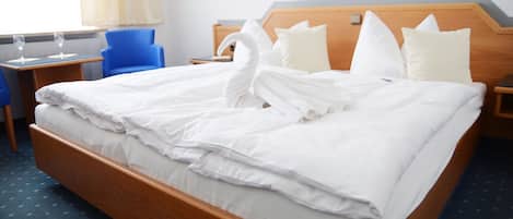Cama bem arrumada com lençóis brancos e uma toalha em formato de cisne, uma cadeira azul, mesa de cabeceira com um copo d'água e mesa de apoio com abajur.