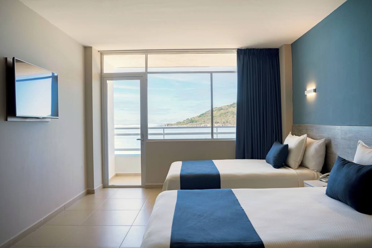 Quarto de hotel com duas camas, televisão fixada na parede, uma janela grande com vista para o mar e uma mesa de cabeceira com abajur.