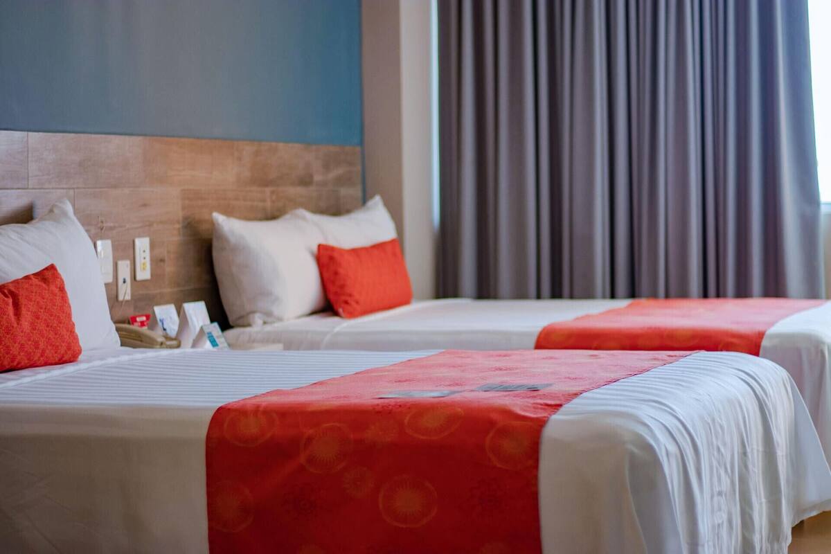 Quarto de hotel com duas camas, cada uma com roupa de cama branca e detalhes vermelhos, uma cabeceira de madeira e vista para uma cortina.