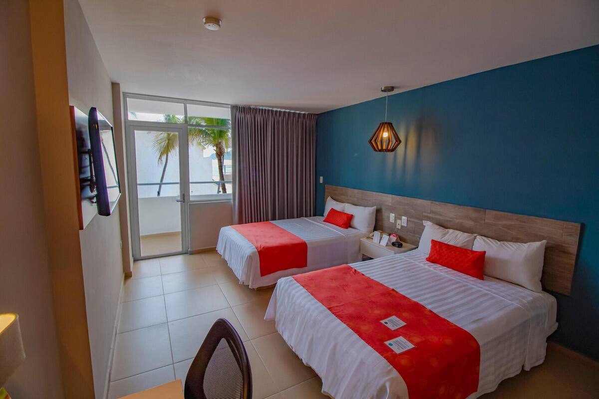 Quarto de hotel com duas camas, uma grande porta de correr de vidro para o exterior, um lustre e uma pequena mesa com uma cadeira.