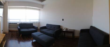 Uma sala de estar com piso de madeira, uma janela grande com cortinas leves e uma TV de tela plana fixada na parede.
