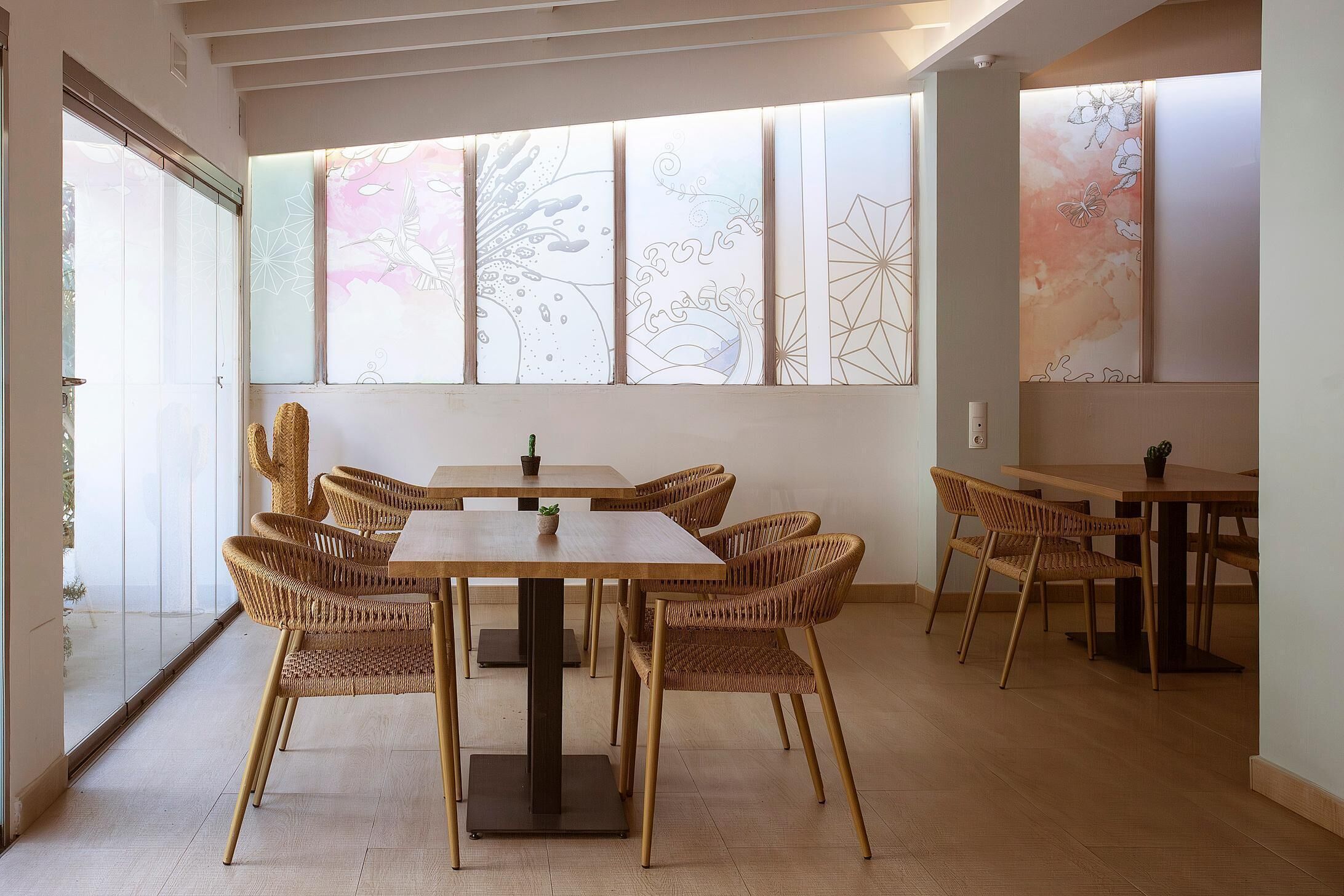 Uma sala de jantar com mesa de madeira e cadeiras de vime, janelas amplas com desenhos decorativos e piso em tons claros.