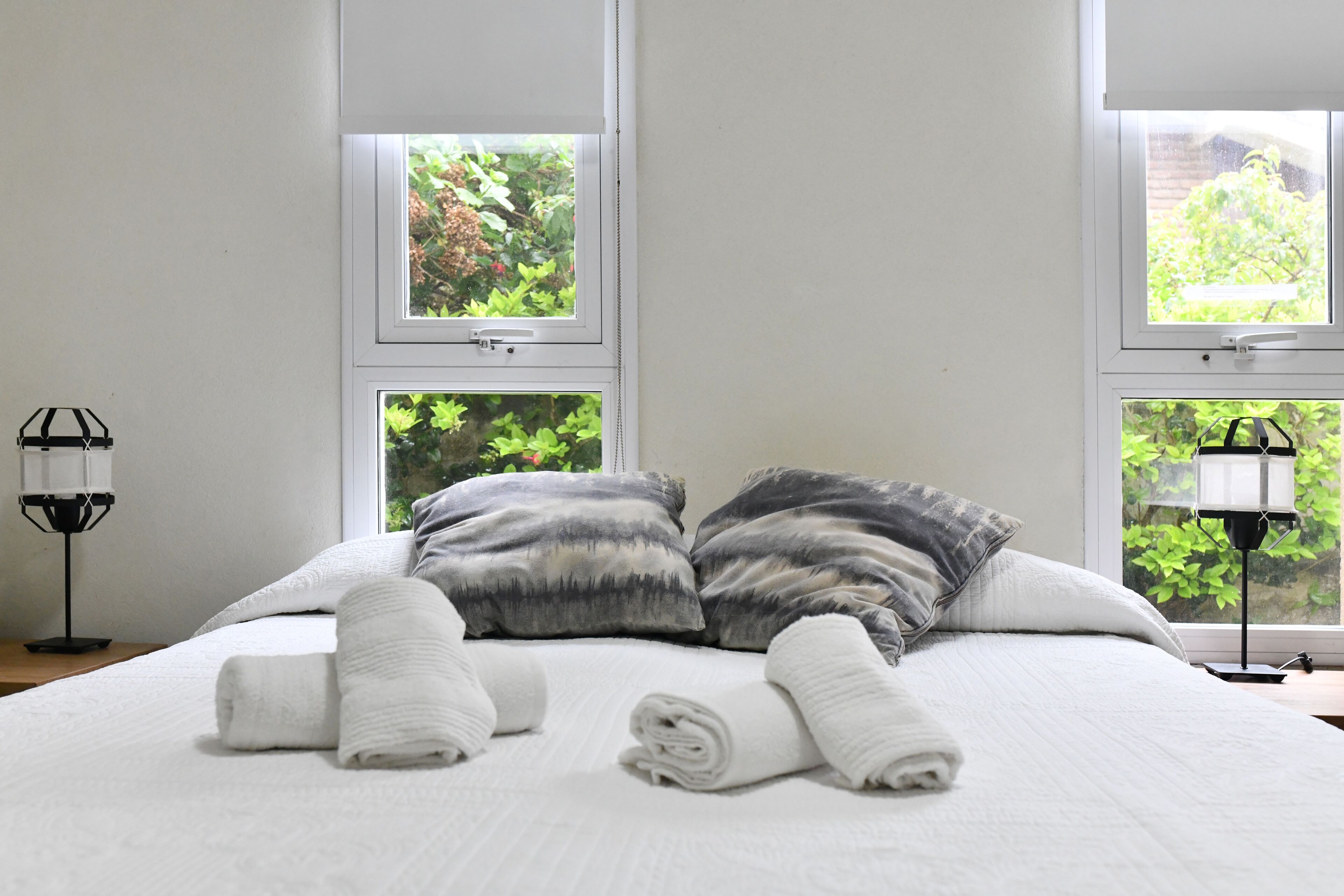 Cama bem arrumada com lençóis brancos e duas almofadas, duas janelas com vista para o verde e duas luminárias de chão de cada lado.