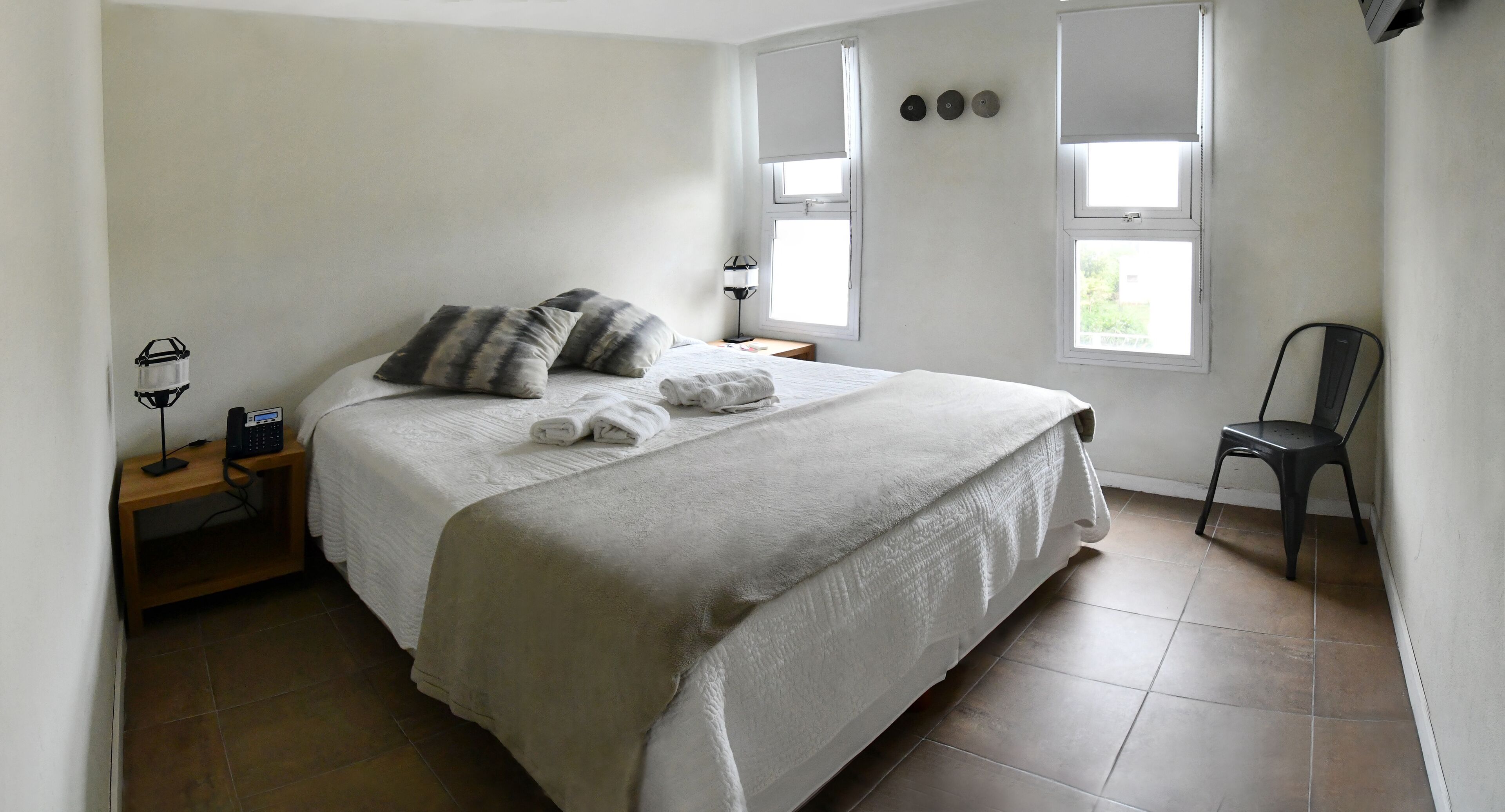 Uma cama bem arrumada com almofadas e toalhas, uma mesa de cabeceira com abajur, e uma cadeira em um quarto com grandes janelas e piso de azulejos.