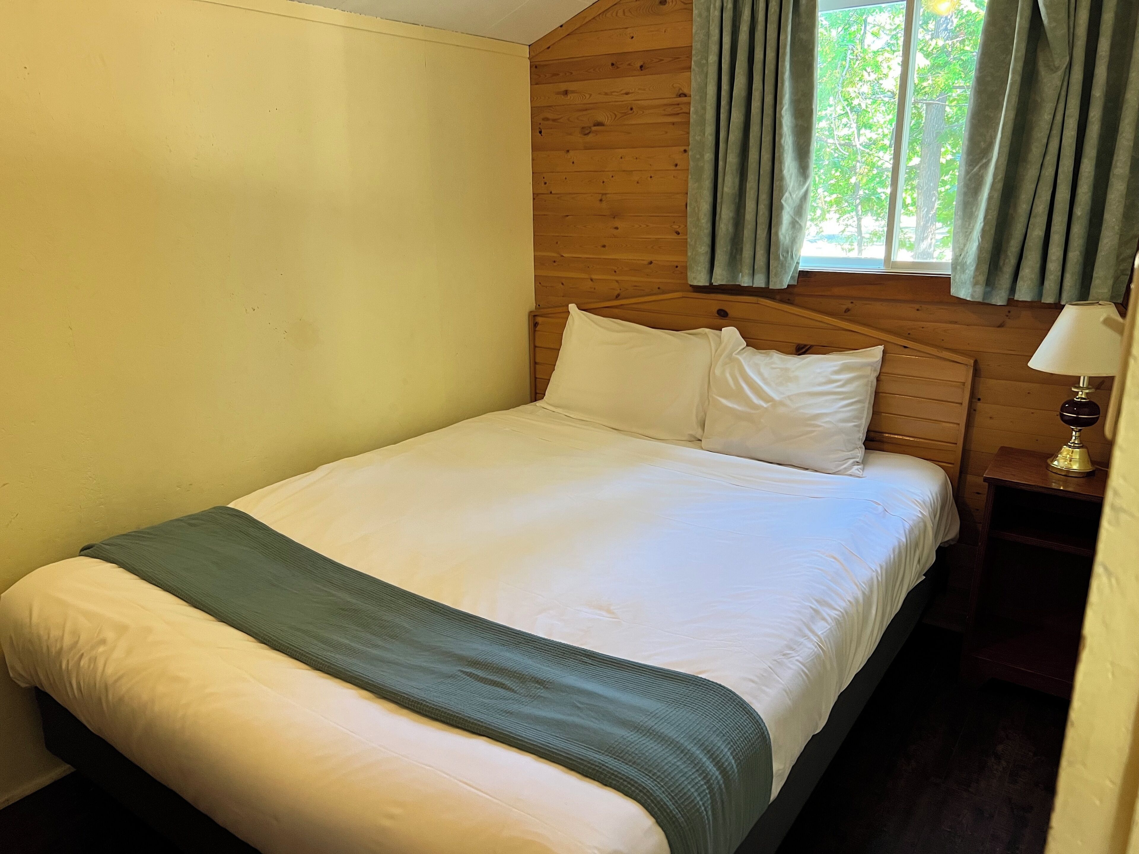 Cama de solteiro com roupa de cama branca e manta verde, cabeceira de madeira, mesa de cabeceira com abajur e janela com cortinas.