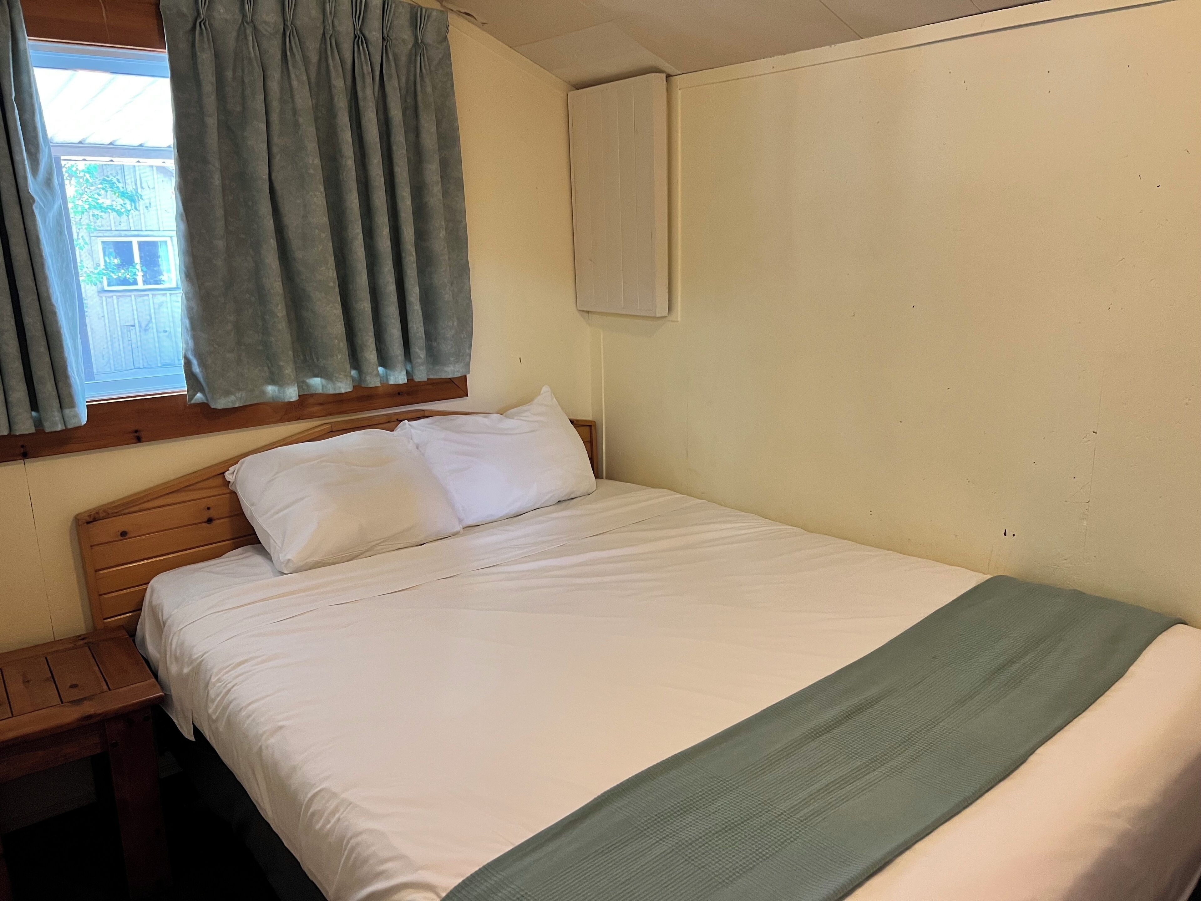 Uma cama de solteiro com cabeceira de madeira, manta verde e janela com cortinas cinzentas.