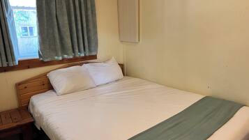 Uma cama de solteiro com cabeceira de madeira, manta verde e janela com cortinas cinzentas.