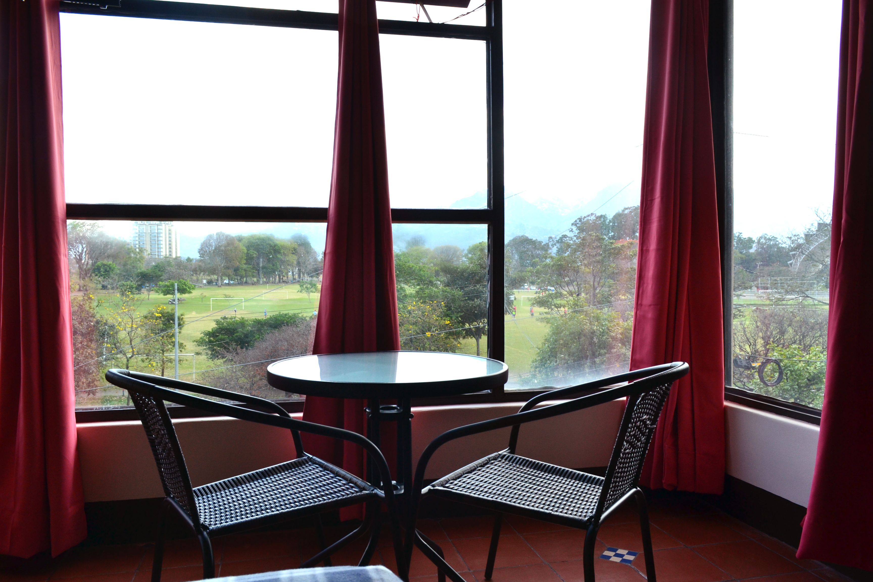 Quarto com mesa redonda e duas cadeiras, uma janela grande com cortinas vermelhas, e vista para um parque e edifícios ao longe.