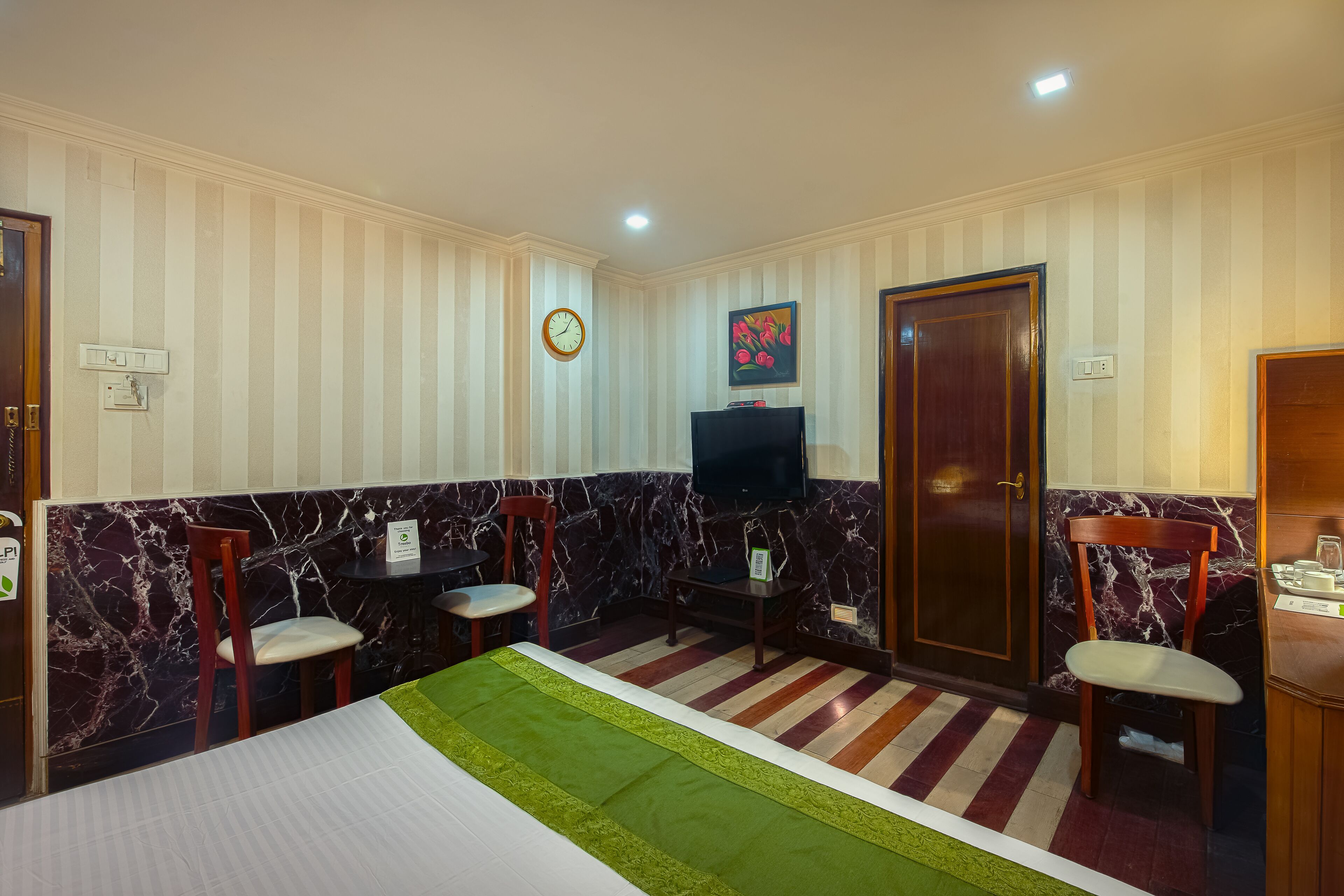 Quarto de hotel com papel de parede listrado, uma cama, uma televisão, um relógio e duas cadeiras.