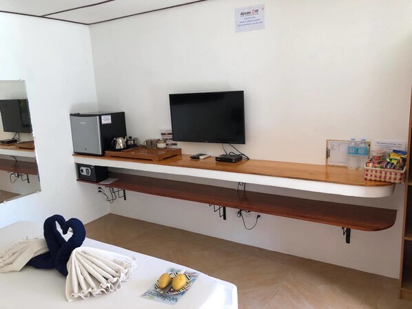 Quarto de hotel com escrivaninha de madeira, TV de tela plana, mini-geladeira, chaleira, bandeja, cesta de petiscos e bebidas, tapete de boas-vindas e arranjo de toalhas.