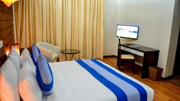 Quarto de hotel com cama, escrivaninha, cadeira, mesa pequena e televisão.
