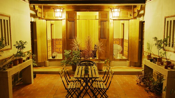 Um pátio externo com mesa e cadeiras de madeira, rodeado por plantas em vasos e piso de azulejos.