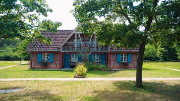 Uma casa tradicional de dois andares com telhado inclinado, rodeada por um gramado bem cuidado e uma grande árvore em primeiro plano.