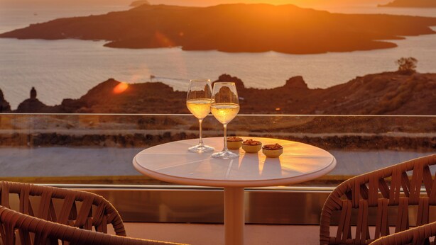 Uma mesa com dois copos de vinho branco e três pequenas tigelas de comida, com vista para o mar ao pôr do sol.