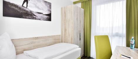 Quarto de hotel com cama, uma cadeira verde, mesa com uma garrafa e um quadro de um surfista.