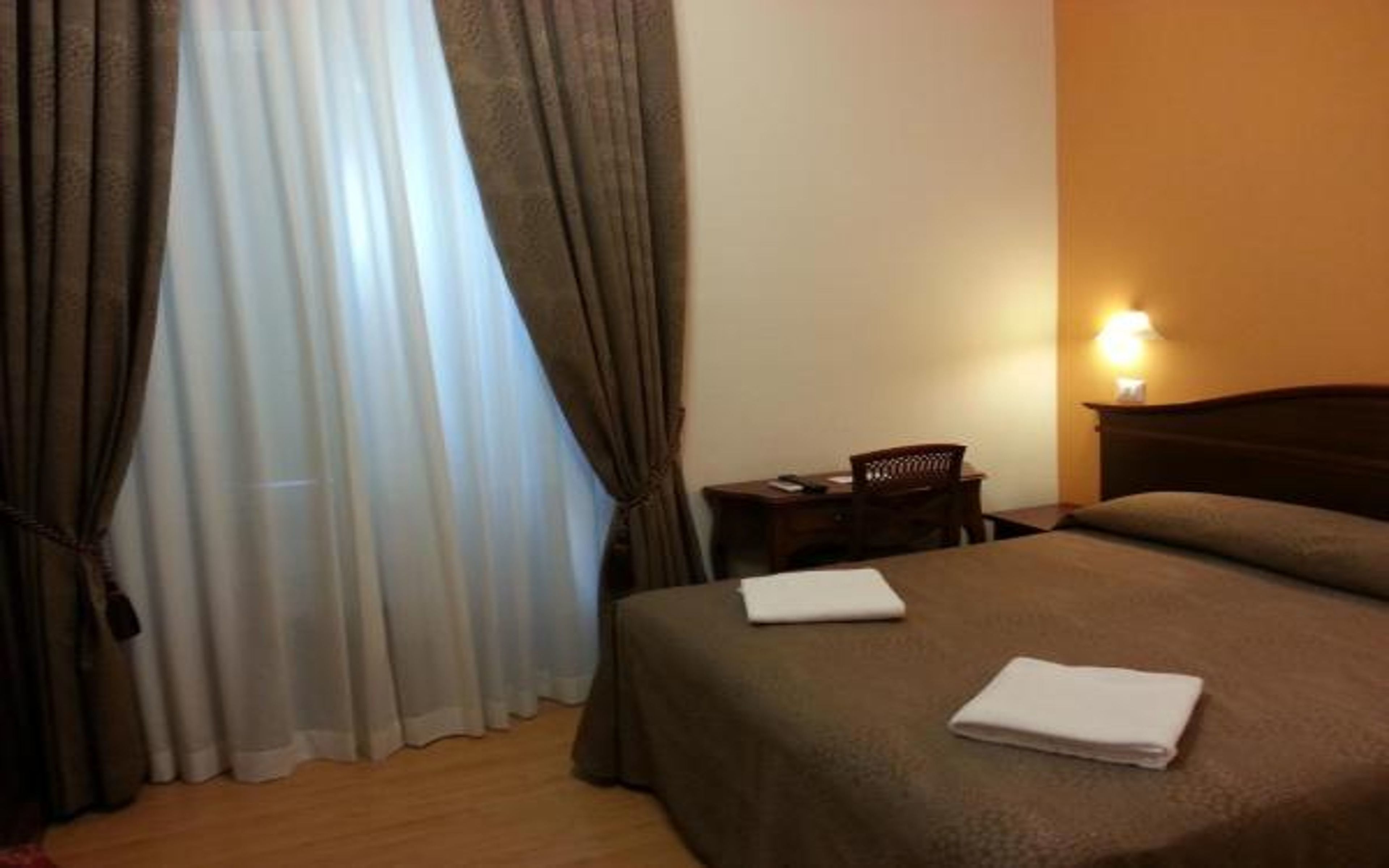 Quarto de hotel com cama, mesa de cabeceira, abajur e cortinas.