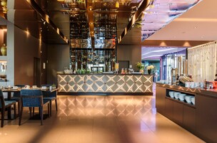 Uma área de bar moderna com balcão de padrão geométrico, um espaço de refeições com cadeiras e mesa, e diversos frascos e copos em prateleiras.