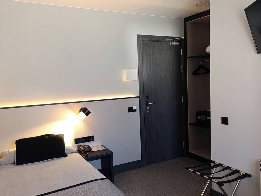 Quarto de hotel com cama, mesa de cabeceira, luminária, guarda-roupa e televisão fixada na parede.