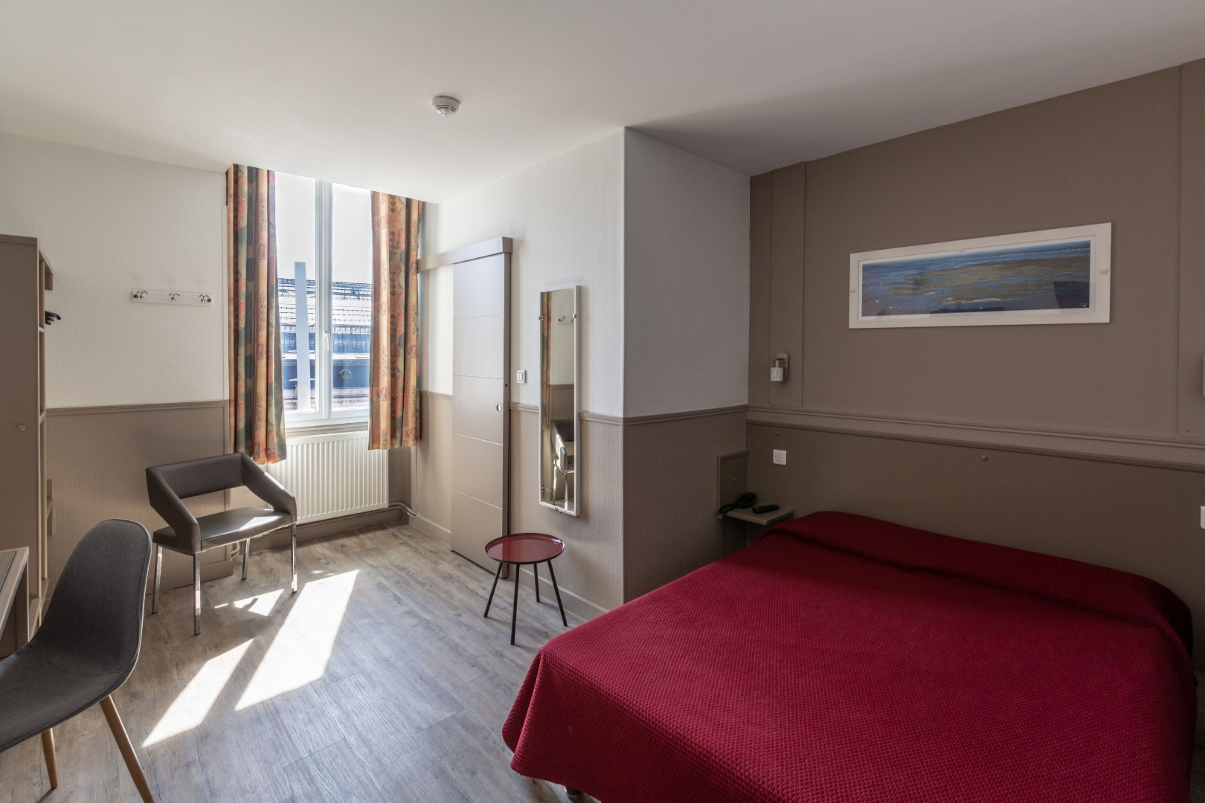Quarto de hotel com cama, escrivaninha, cadeira, mesinha e janela com cortinas.
