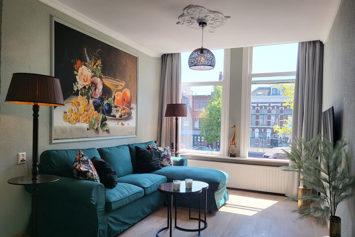 Uma sala de estar com um sofá verde-água, uma mesa de centro redonda, um quadro de frutas, uma estatueta de girafa e vista de prédios através de grandes janelas.