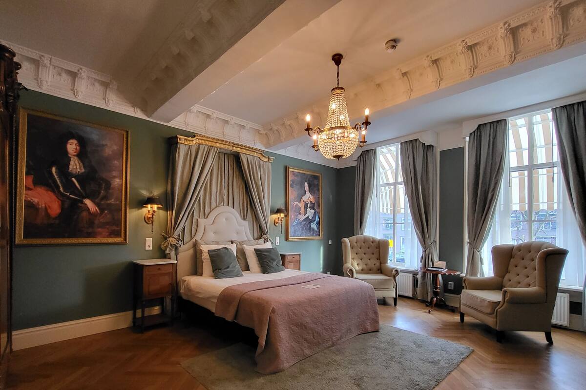 Um quarto espaçoso com uma cama grande, duas poltronas, um candelabro e obras de arte nas paredes.