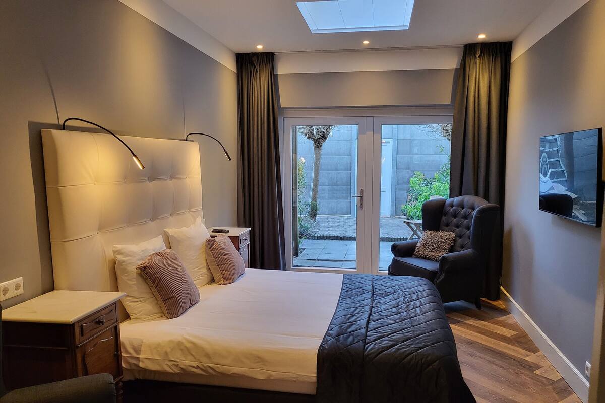 Quarto de hotel moderno com uma cama grande, uma escrivaninha, uma cadeira, uma televisão e vista para o exterior.