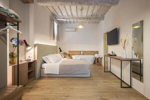 Quarto moderno com piso de madeira, cama com roupa de cama branca, mesa de cabeceira, espelho e TV fixada na parede.